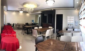 Uma sala de jantar com várias mesas e cadeiras, um lustre e uma área de cozinha com pia e janela.