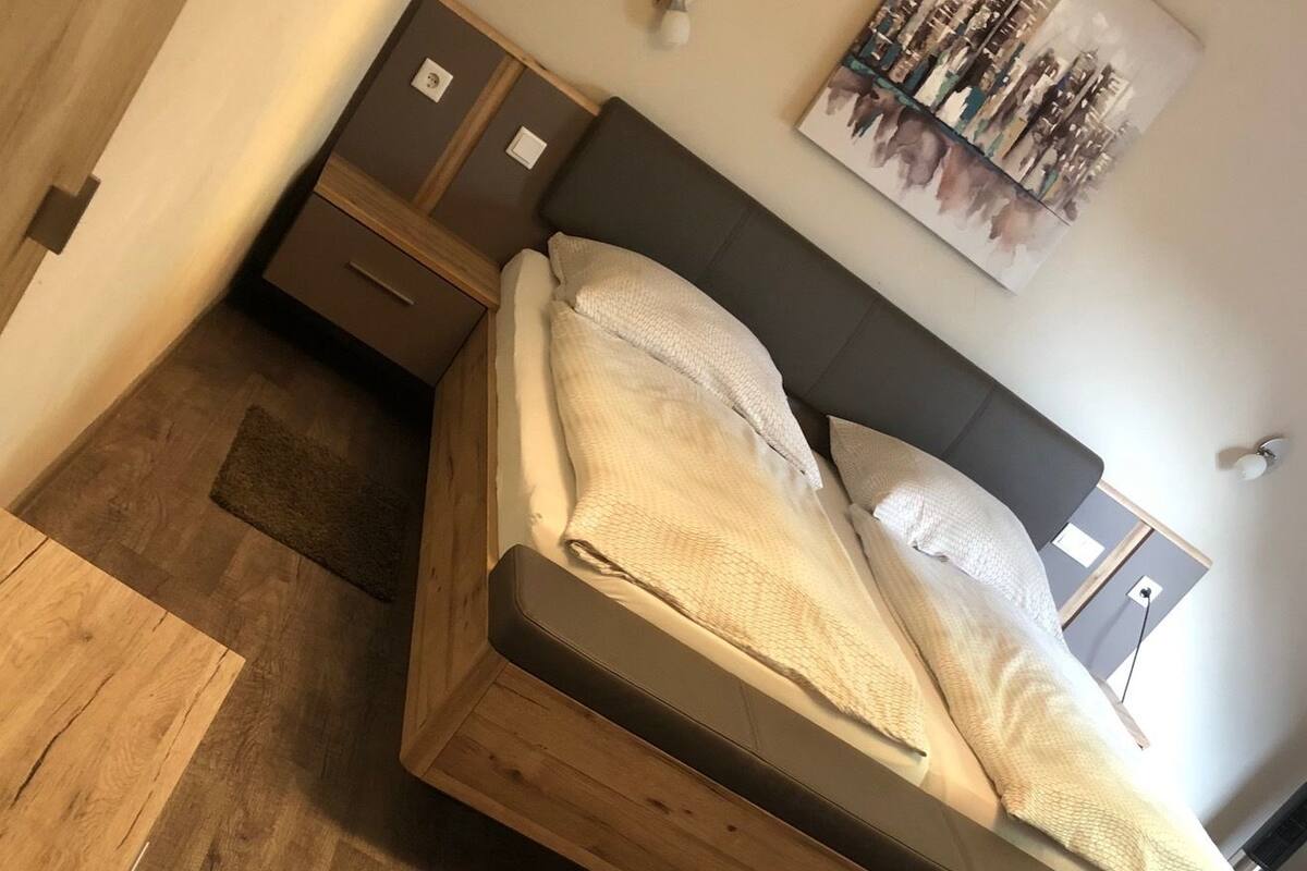 Um quarto moderno com estrutura de cama de madeira, cabeceira escura e uma pintura de paisagem urbana na parede.