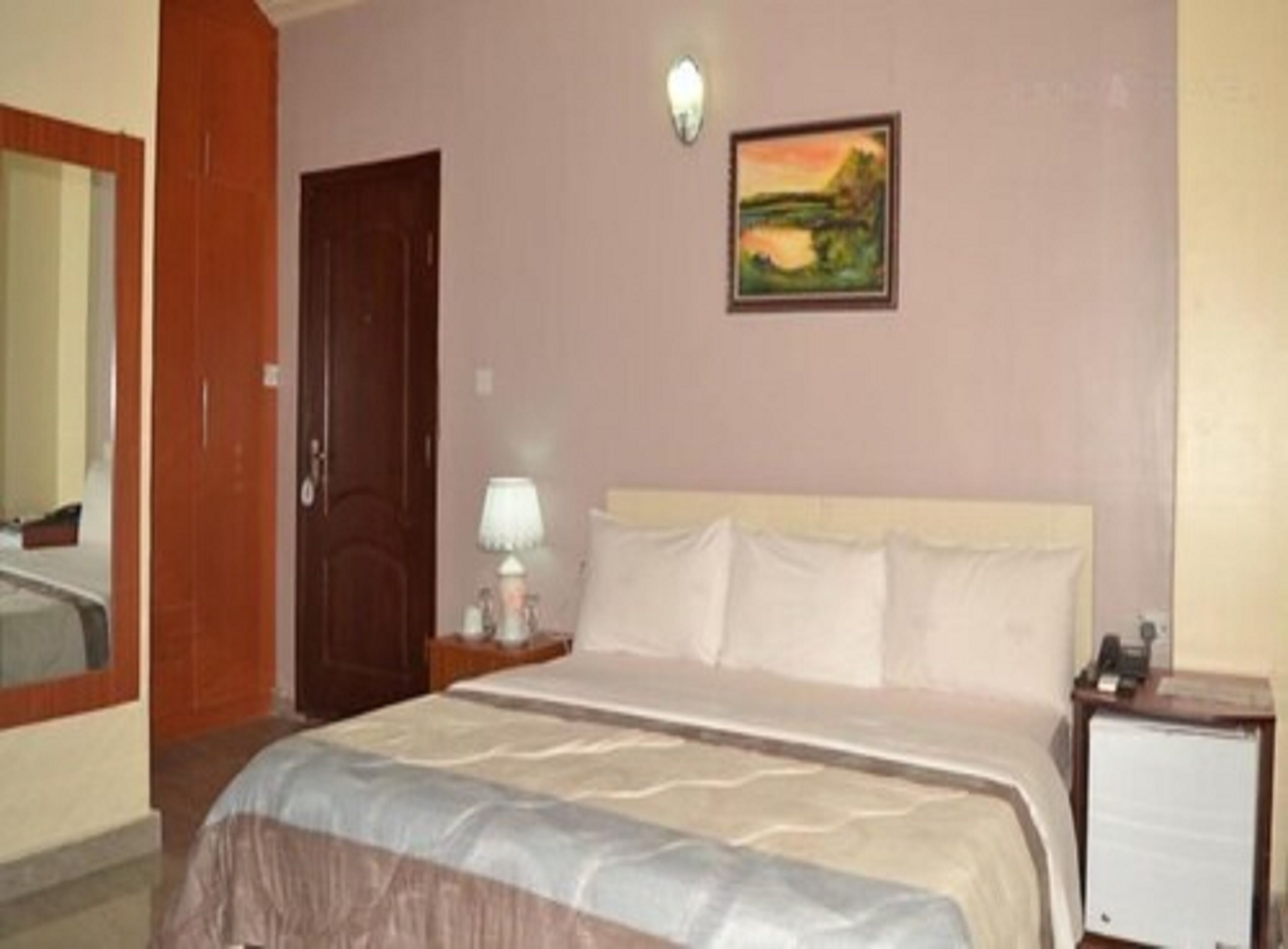 Quarto de hotel com uma cama, mesa de cabeceira com abajur, quadro na parede e uma porta.