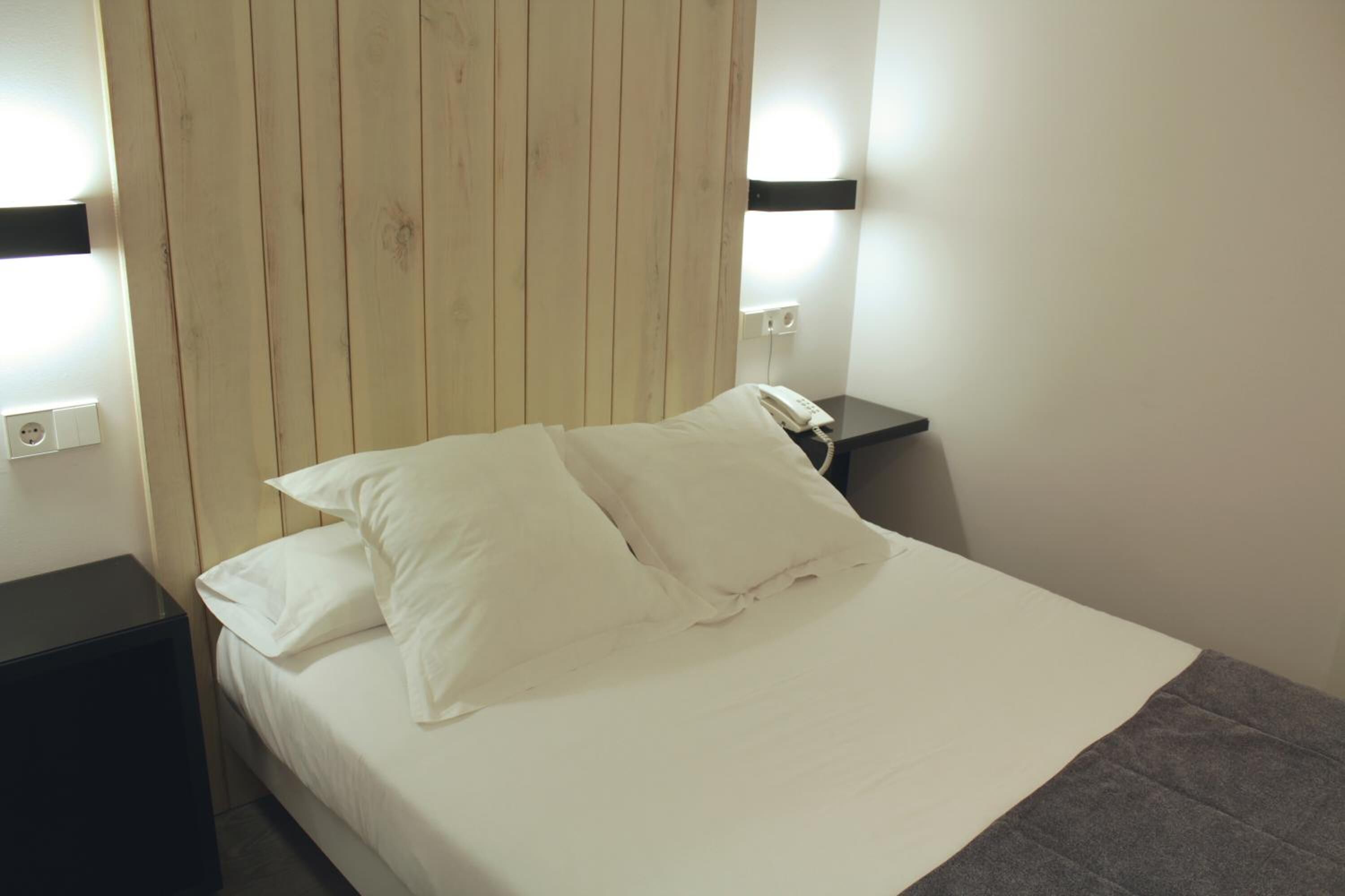 Cama bem arrumada com roupa de cama e almofadas brancas, um telefone ao lado da cama e cabeceira de madeira.