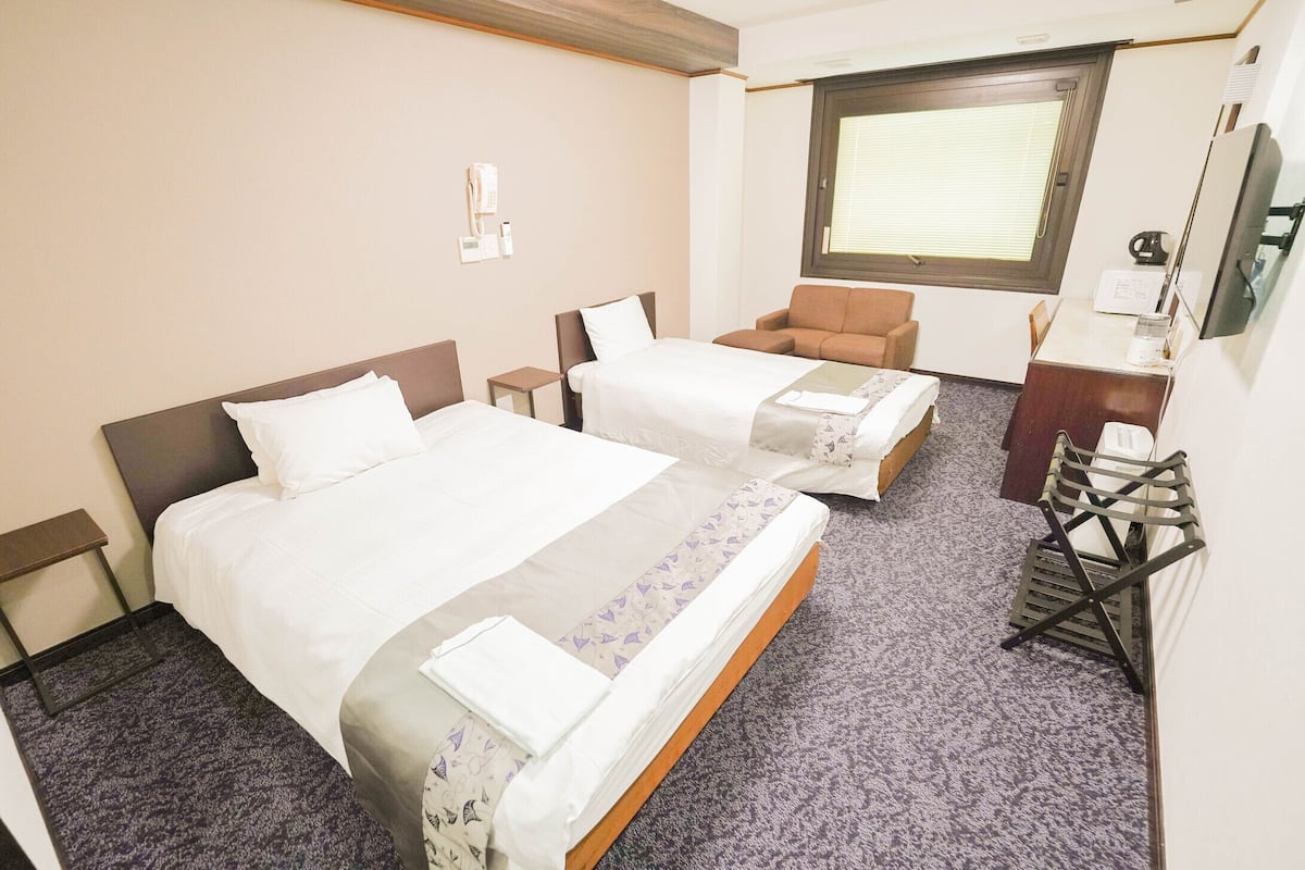 Quarto de hotel com duas camas, um sofá, uma escrivaninha, uma cadeira, uma janela e um espelho.
