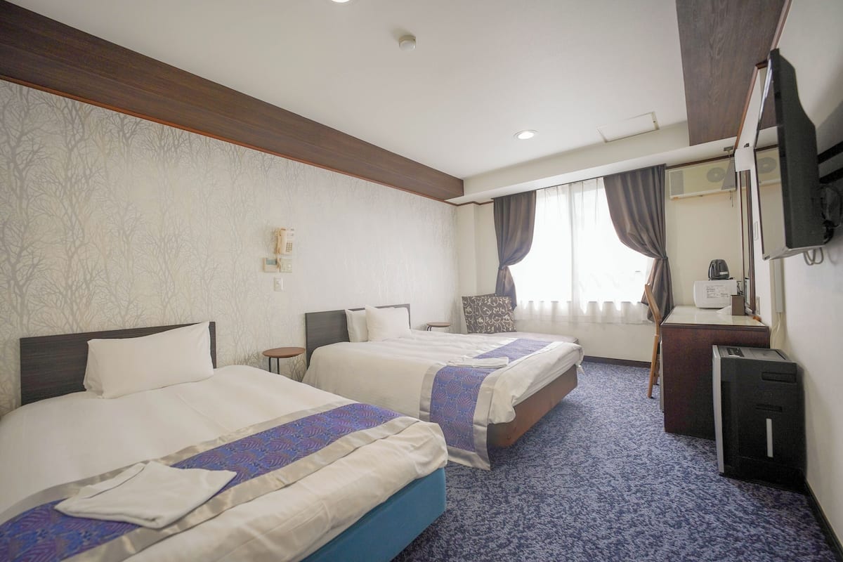 Quarto de hotel com duas camas, uma escrivaninha, uma cadeira, uma televisão e uma janela com cortinas.