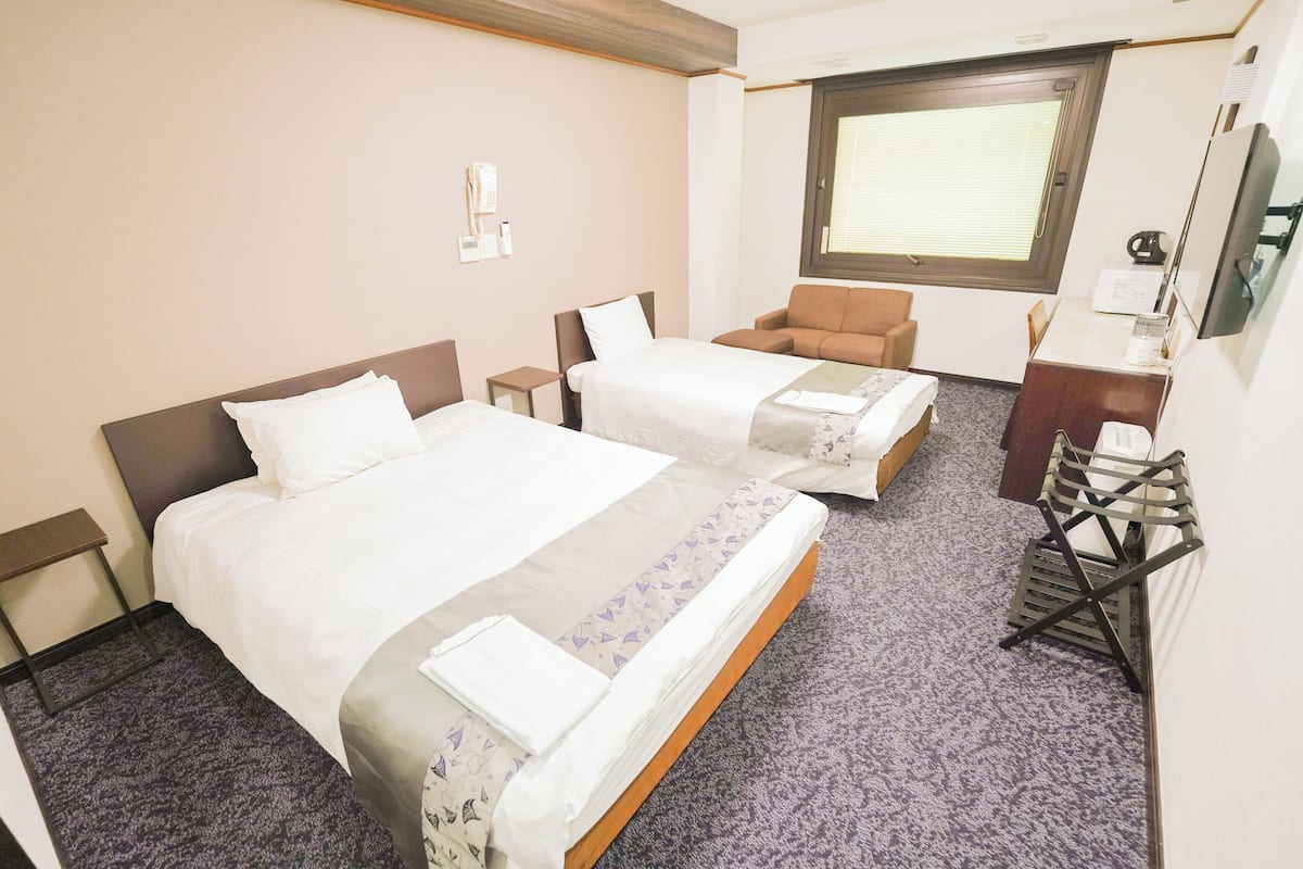 Quarto de hotel com duas camas, uma escrivaninha, uma cadeira, uma janela com persianas e um telefone.