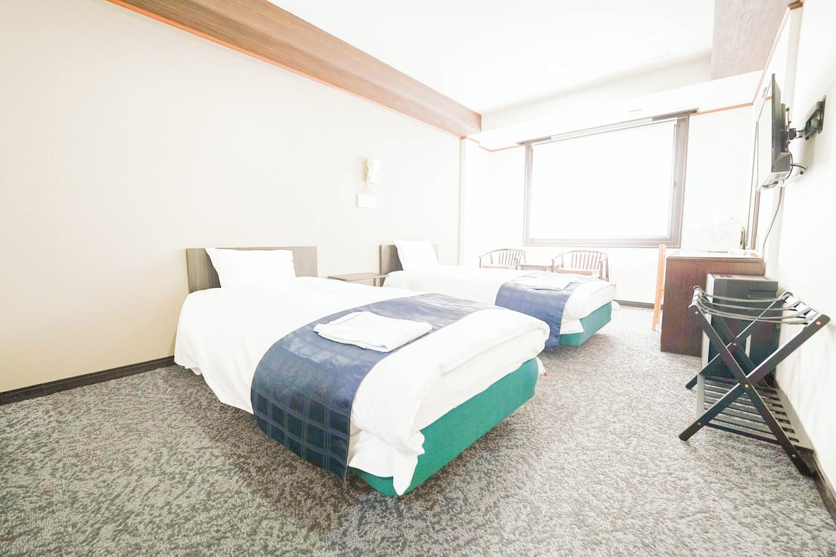 Quarto de hotel com duas camas, uma escrivaninha, uma cadeira e uma janela grande.