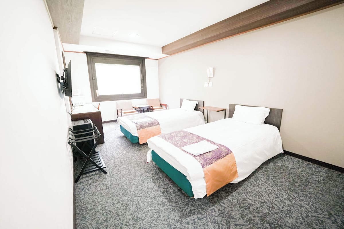 Quarto de hotel com duas camas, uma escrivaninha, uma cadeira, uma televisão e uma janela.