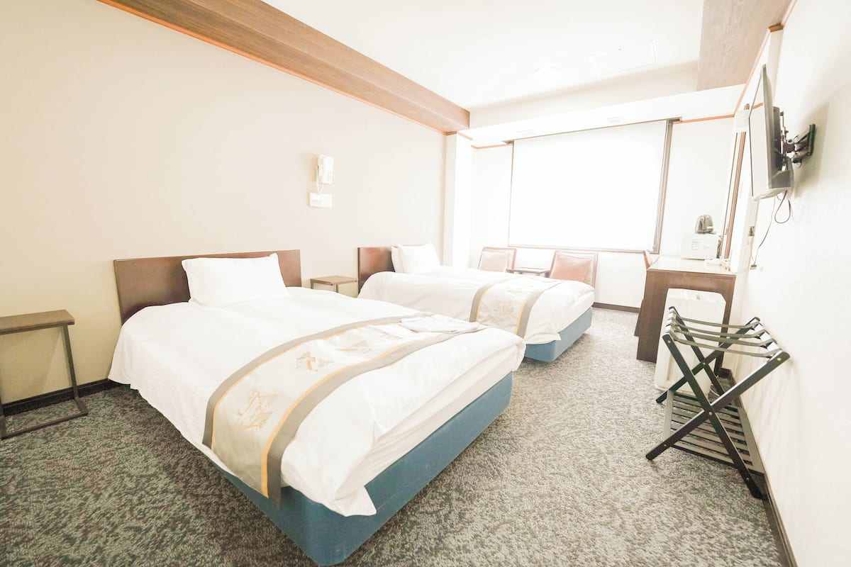Quarto de hotel com duas camas, uma mesinha, uma cadeira, uma escrivaninha e uma janela.