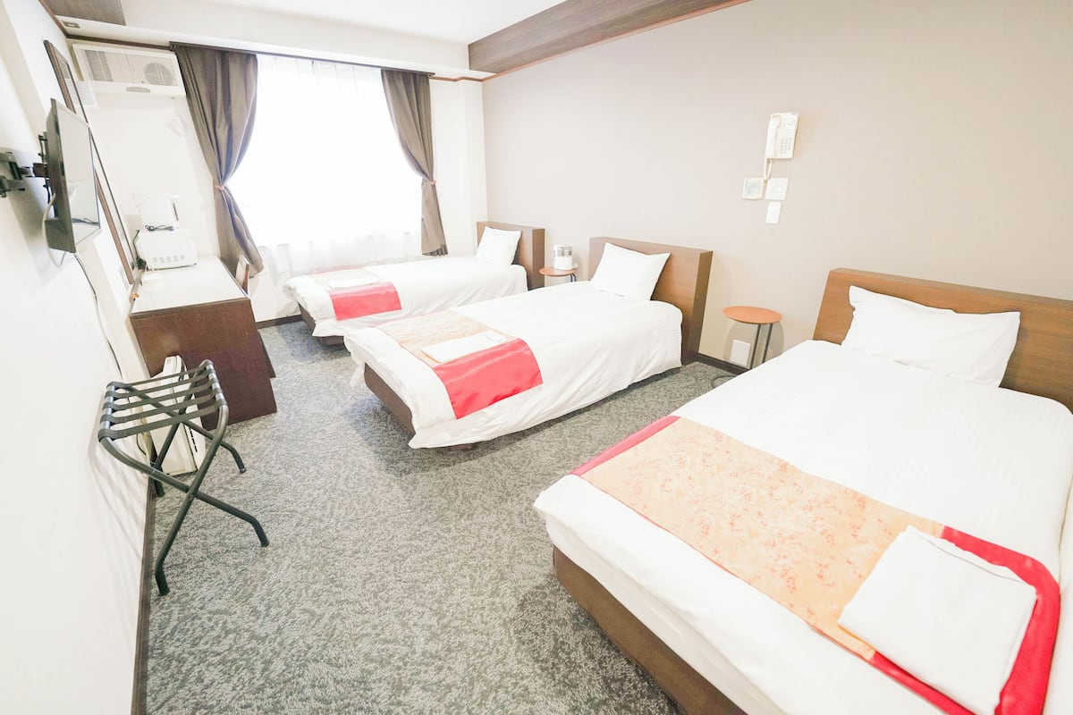 Quarto de hotel com três camas, uma escrivaninha, uma cadeira, um telefone e uma janela com cortinas.