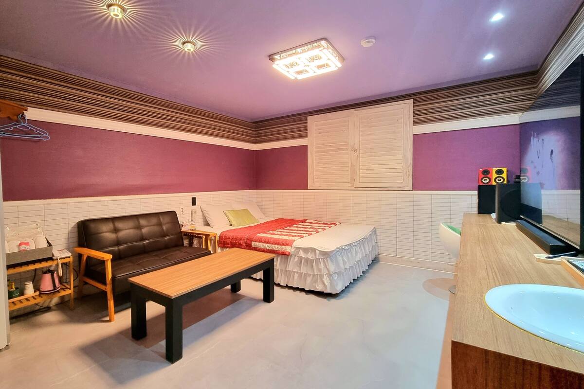 Quarto com cama, sofá, mesa pequena, televisão e área de banheiro.