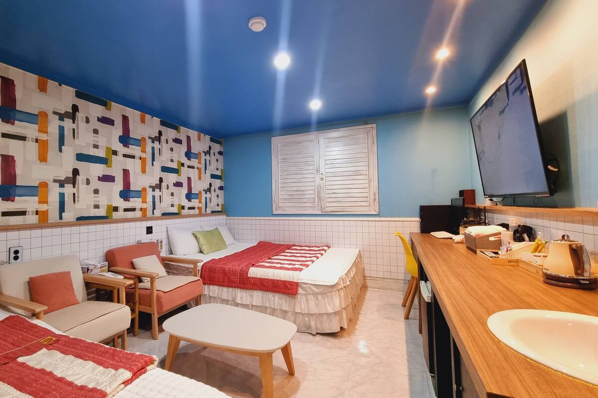 Quarto de hotel com duas camas, um espaço de estar pequeno, uma escrivaninha de madeira, uma pia e uma televisão.