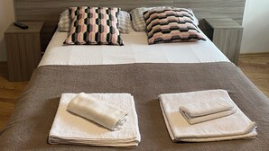 Cama bem arrumada com cabeceira marrom, duas almofadas com estampas geométricas e duas toalhas dobradas sobre um espalha. 