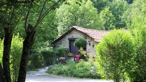 Uma casa de pedra rústica com telhado de telhas vermelhas, rodeada por vegetação exuberante e um caminho de cascalho que leva à entrada.