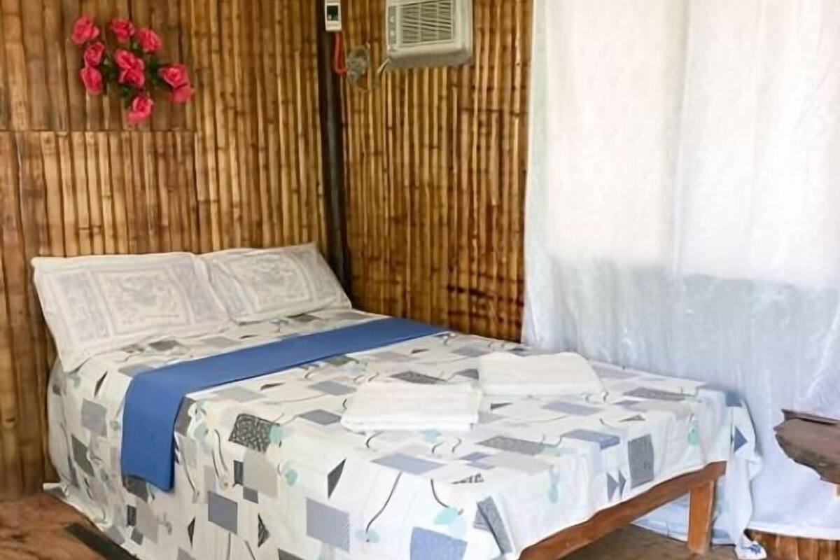 Uma cama de solteiro com colcha estampada, painéis de parede de madeira, um pequeno aparelho de ar condicionado e uma janela com cortinas brancas.