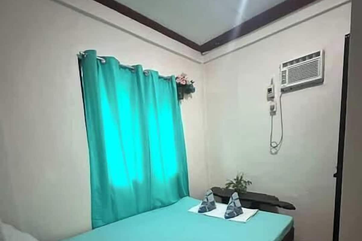 Quarto pequeno com cama de solteiro, colcha verde-água, travesseiro branco e uma mesinha com uma planta.