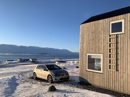 Uma pequena cabana de madeira com um carro estacionado em frente, localizada perto de uma paisagem nevada com um corpo d'água e montanhas ao fundo.
