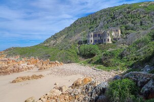 Um edifício histórico de pedra com uma torre, localizado numa encosta rochosa com uma praia ao primeiro plano.