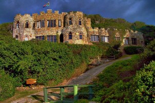 Um edifício imponente, semelhante a um castelo de pedra, com várias janelas e uma bandeira, rodeado por vegetação exuberante e um caminho.