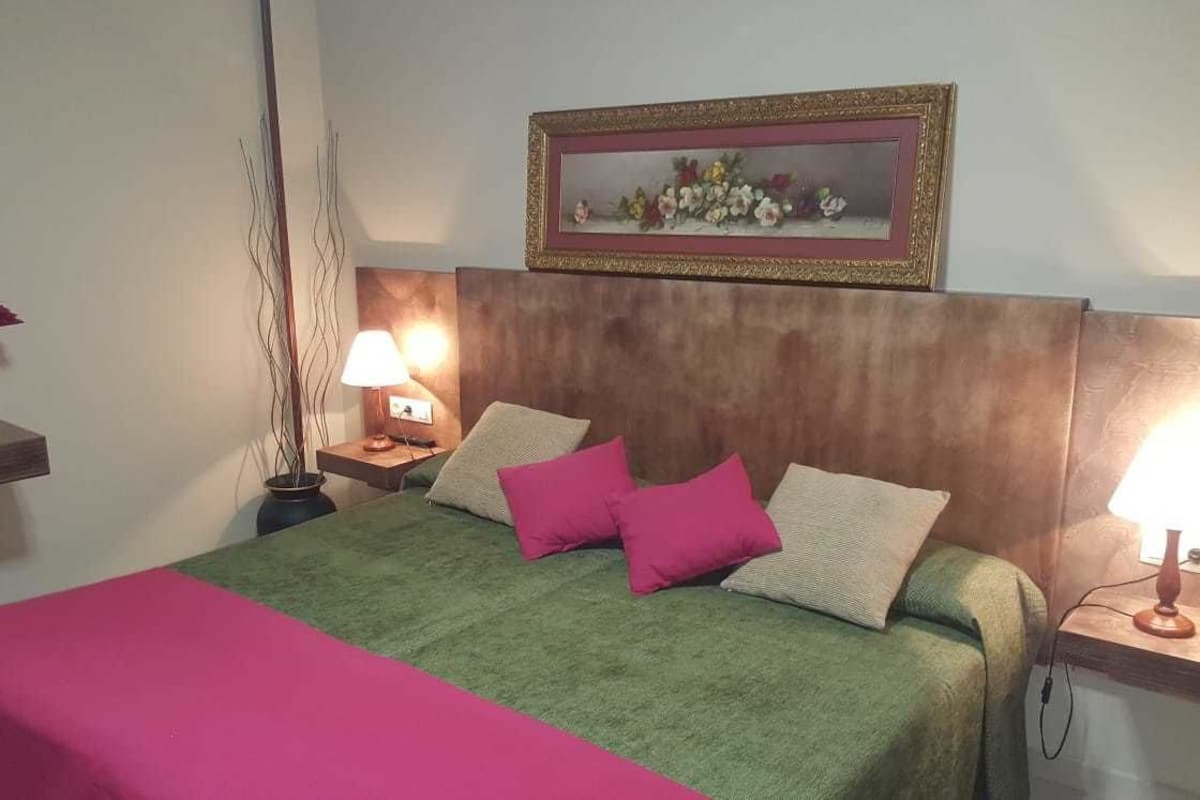 Uma cama bem arrumada com um edredom verde e duas almofadas, uma mesinha de cabeceira com uma luminária, um quadro emoldurado acima da cama e um vaso com flores em uma prateleira.