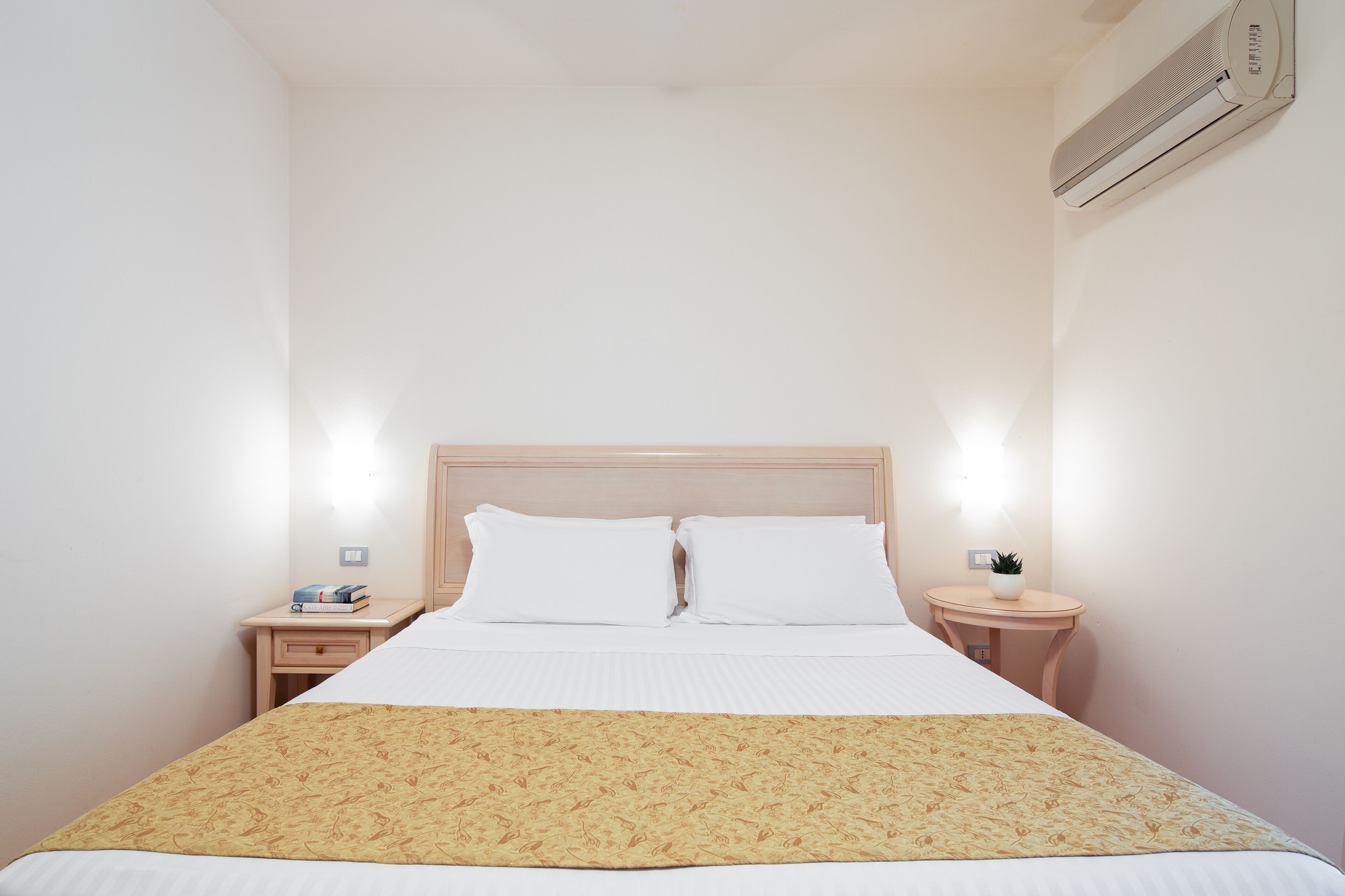 Cama bem arrumada com lençóis brancos e manta estampada, duas mesas de cabeceira com abajures, e ar condicionado fixado na parede.