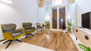 Quarto espaçoso com piso de madeira, dois poltronas estampadas, uma mesa pequena e uma televisão sobre um móvel.