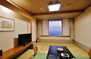 Quarto com televisão sobre suporte de madeira, mesa com tapete preto e vista para o mar através de uma janela com painéis shoji.