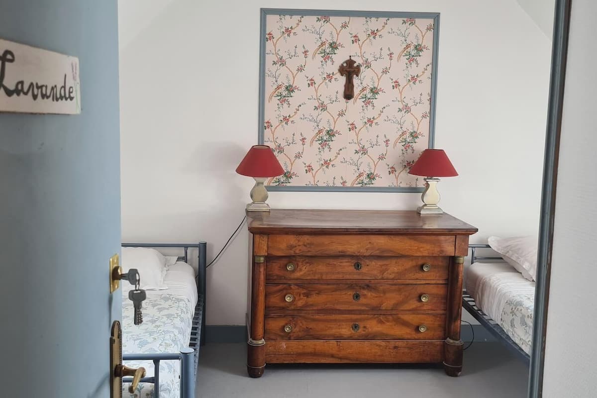 Quarto com cómoda de madeira, duas luminárias, cama com roupa de cama floral e parede com papel de parede floral e uma cruz.