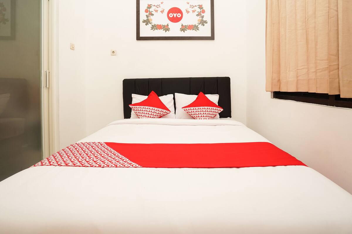 Uma cama com colcha branca e vermelha, duas almofadas vermelhas e um quadro na parede.
