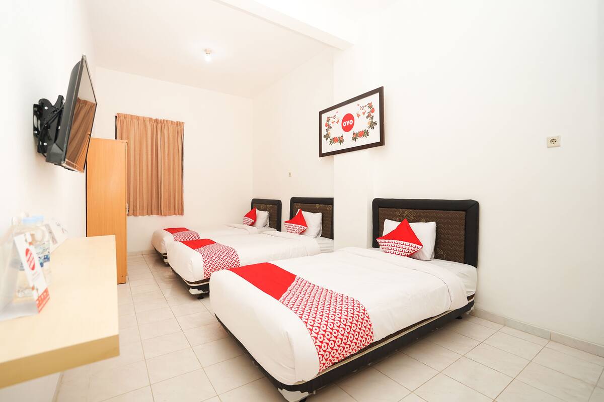 Quarto de hotel com três camas, cada uma com roupa de cama branca e detalhes vermelhos, uma escrivaninha de madeira e um quadro na parede.
