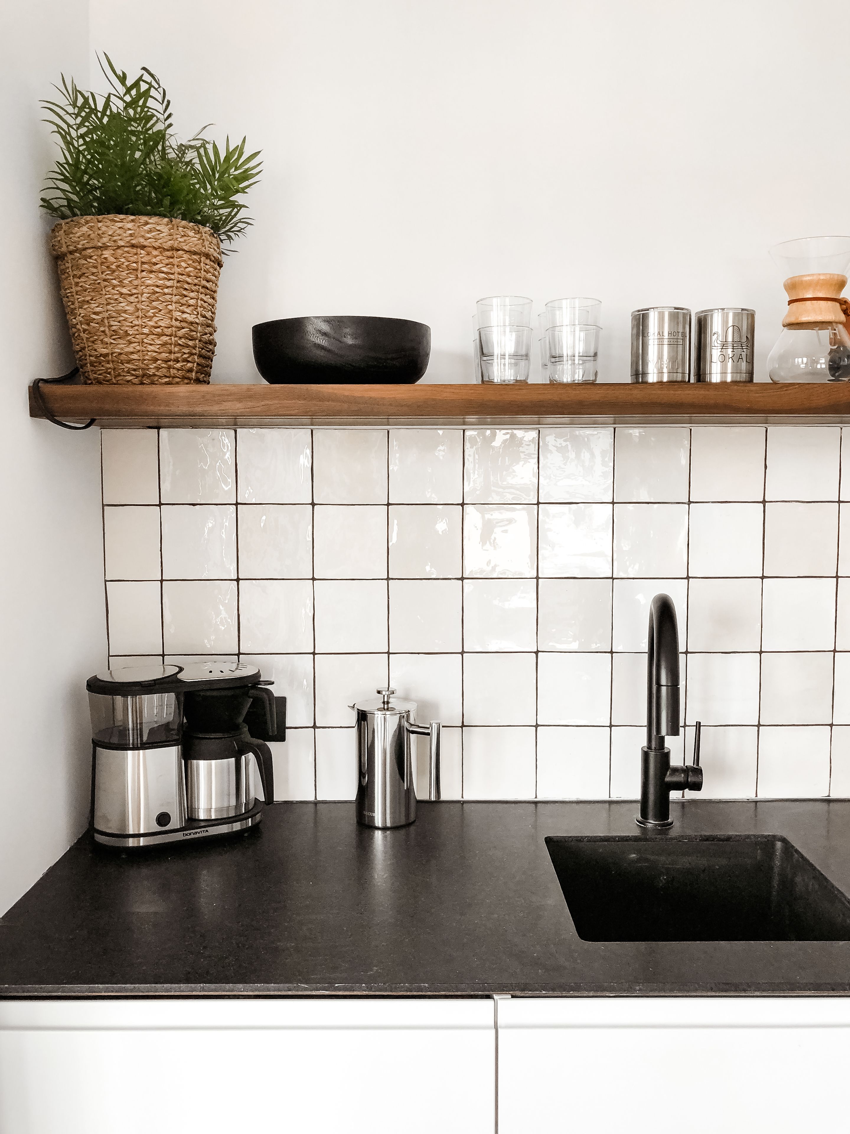Uma cozinha com bancada preta, máquina de café de aço inoxidável, pia preta e painel atrás do fogão revestido com azulejos.