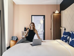 Quarto de hotel moderno com uma cama, um laptop, uma mesa com cadeira, uma mala e um alto-falante fixado na parede.