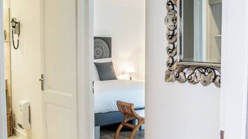 Quarto de hotel com piso de madeira, cama com cabeceira estampada, uma cadeira, espelho com moldura ornamentada e porta para o banheiro.