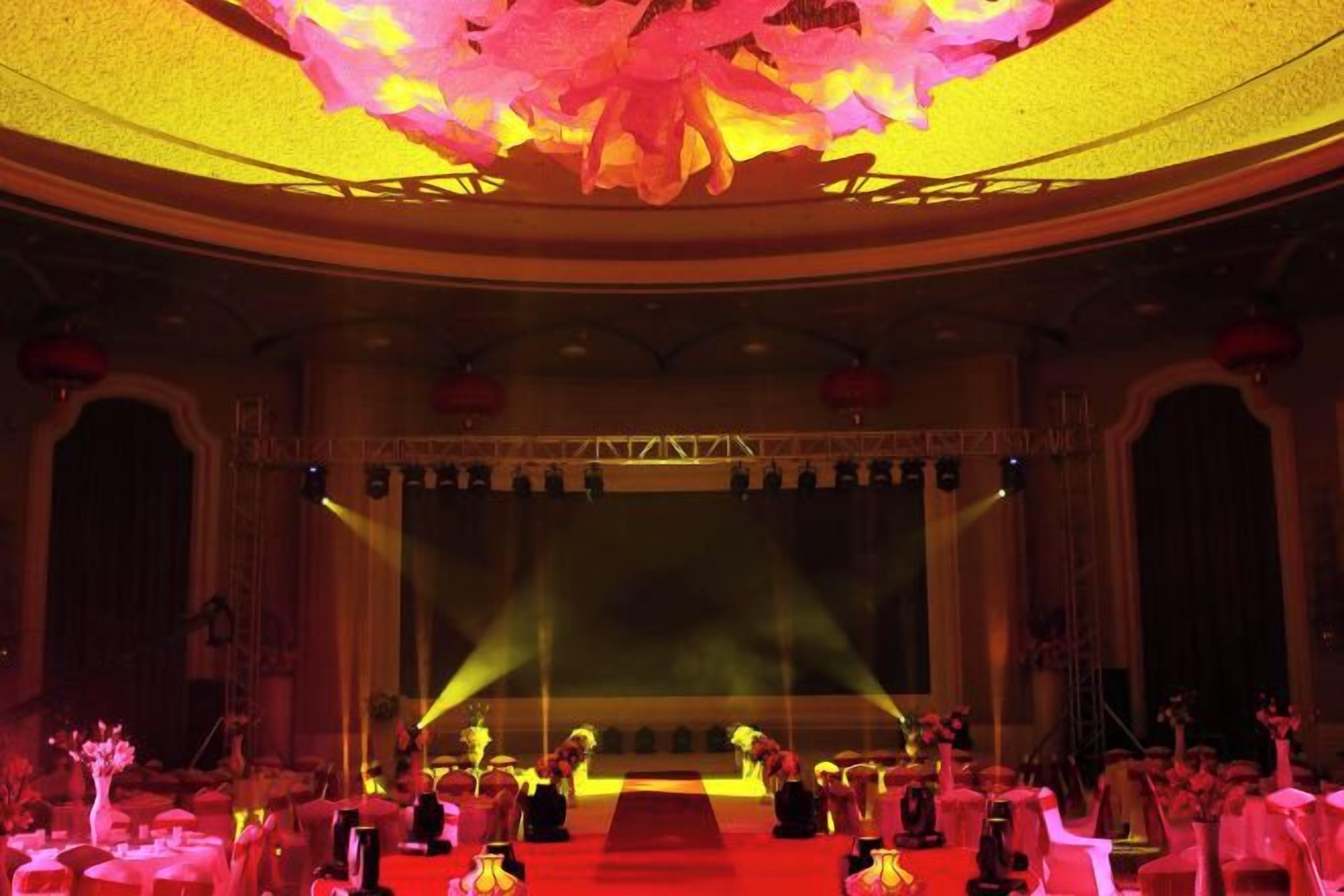 Palco com tapete vermelho, lustre e público sentado em mesas.