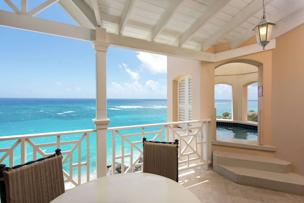 Uma área externa coberta com vista para o oceano, piscina e uma mesa com cadeiras.