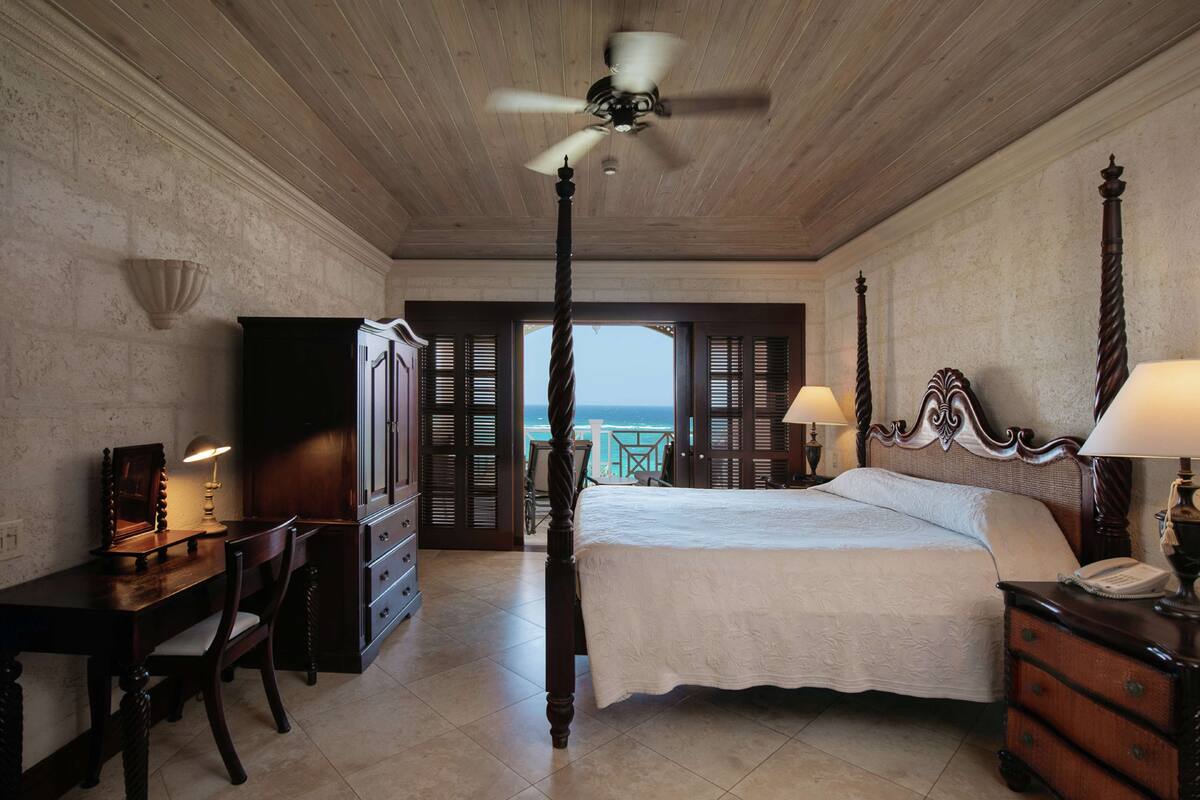 Quarto com cama de dossel, escrivaninha, cadeira, guarda-roupa e vista para o mar por uma porta.
