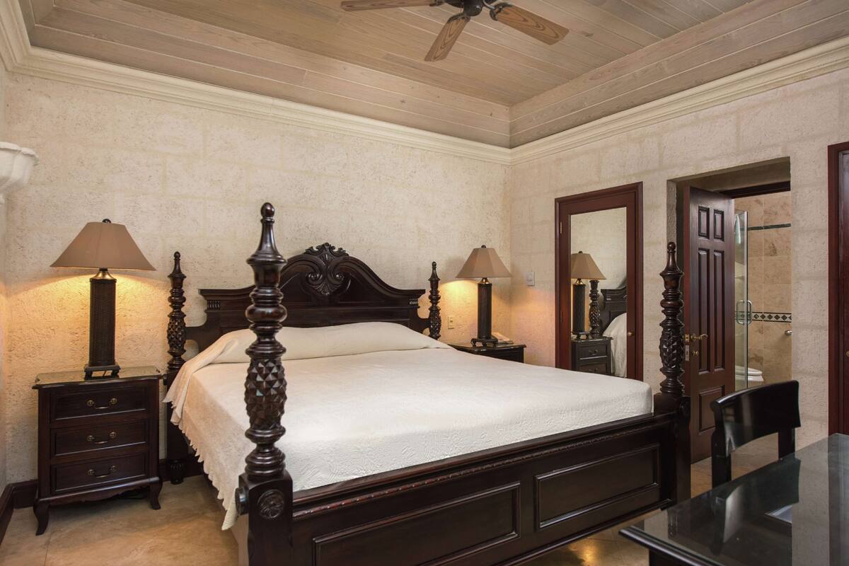 Um quarto com uma cama grande, duas mesas de cabeceira com luminárias, uma cadeira de madeira e um espelho na parede.