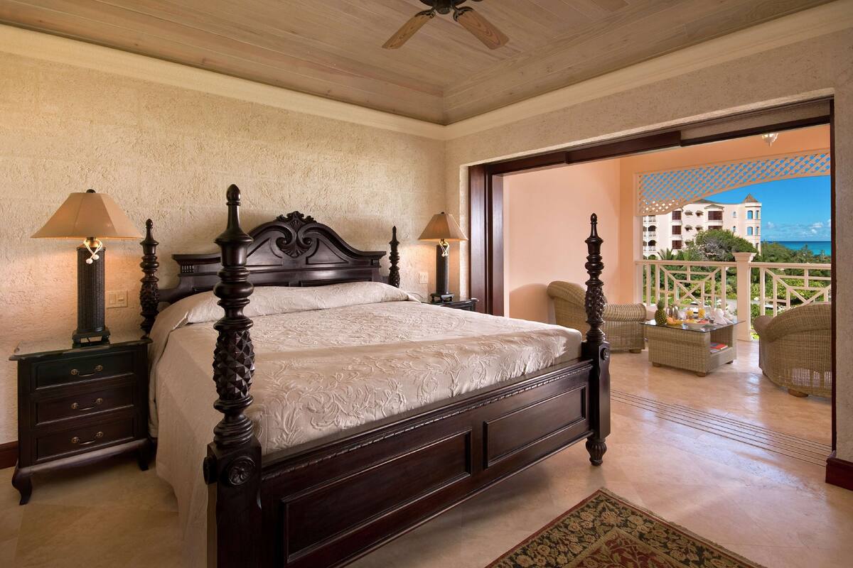 Quarto com cama grande, duas mesas de cabeceira com abajures, ventoinha de teto e vista para o mar por uma porta aberta.