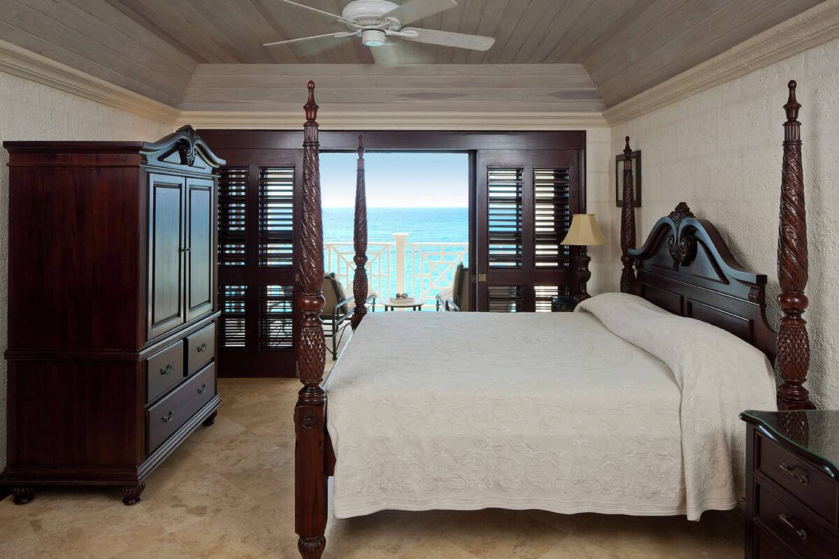 Quarto com cama de dossel, guarda-roupa de madeira e vista para o mar através de uma varanda com persianas.