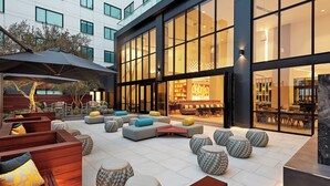 Uma área externa com assentos de madeira, bancos e banquetas redondas, um grande guarda-sol, e um edifício moderno com grandes janelas ao fundo.