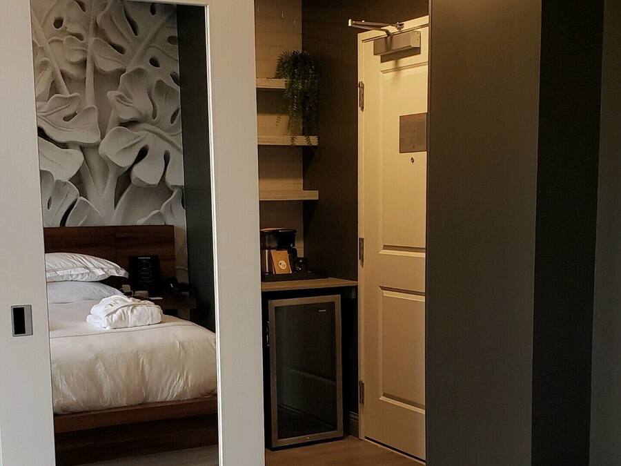 Quarto de hotel moderno com uma cama grande, uma pequena escrivaninha, um espelho e uma porta para o banheiro.