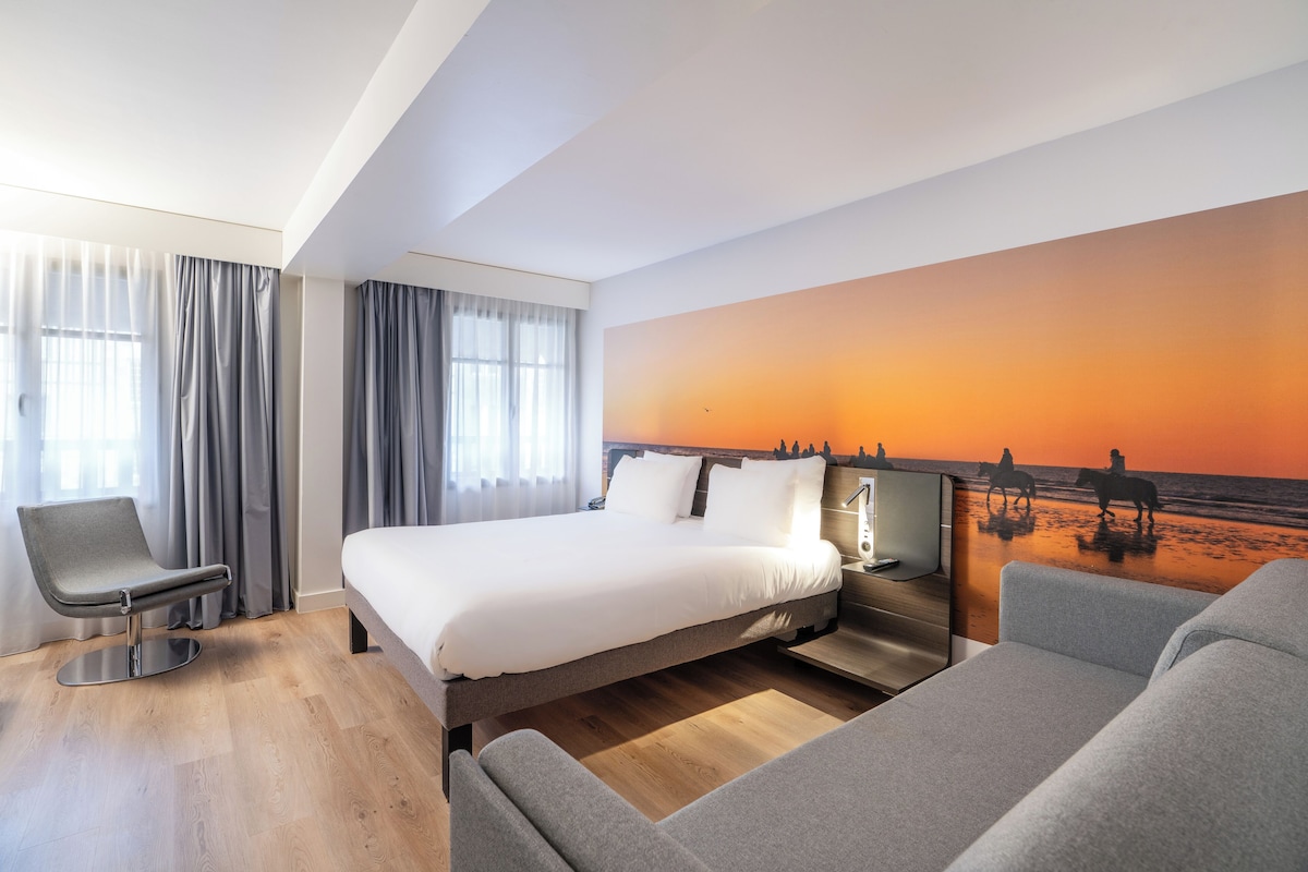 Quarto de hotel com cama, escrivaninha, cadeira e um grande mural na parede retratando um pôr do sol sobre o oceano.