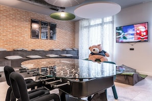 Quarto moderno com mesa de vidro, assentos confortáveis, um urso de pelúcia grande e uma TV exibindo uma cena festiva.