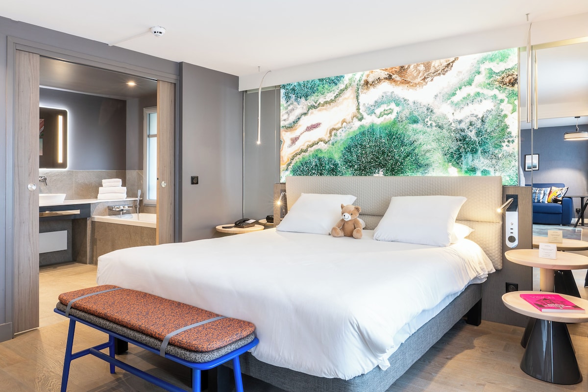 Quarto de hotel moderno com uma cama grande, uma escrivaninha, uma cadeira, uma mesa pequena e uma janela ampla com vista para árvores.