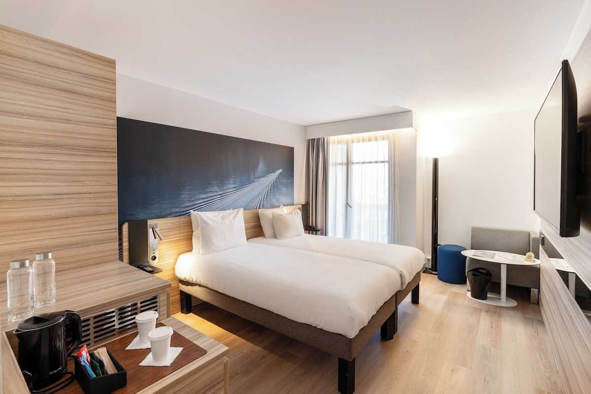 Quarto de hotel moderno com uma cama grande, televisão de tela plana, escrivaninha com cadeira e janela com cortinas.