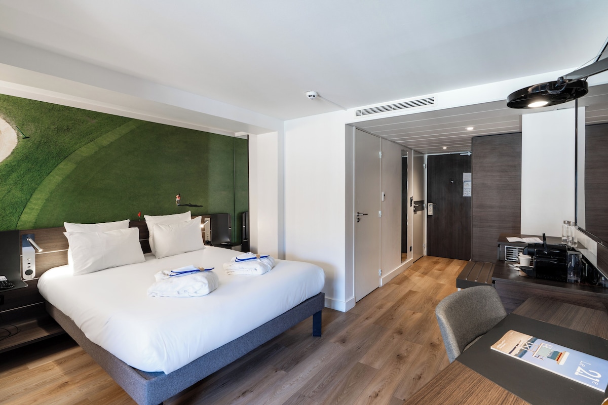 Quarto de hotel moderno com uma cama grande, uma mesa com cadeira, parede verde e porta que dá para outro cômodo.