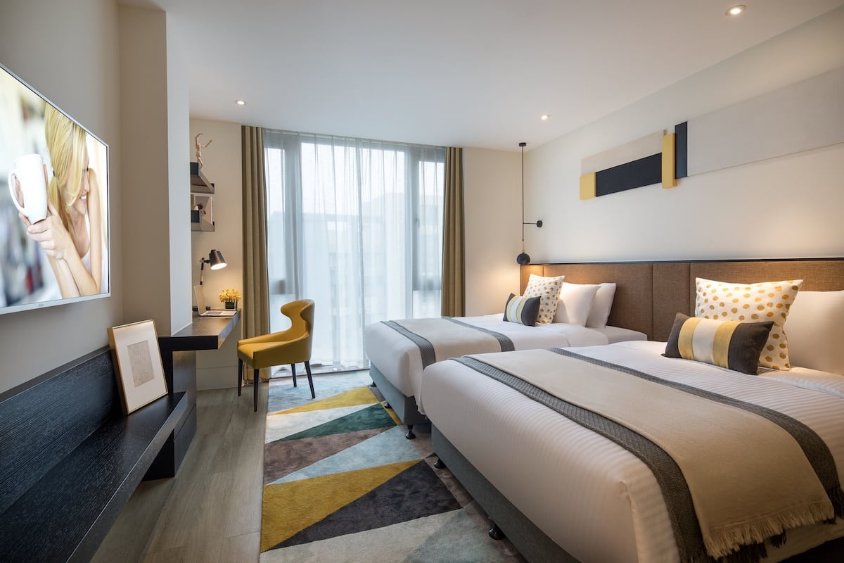 Quarto de hotel com duas camas, uma escrivaninha, uma cadeira e uma janela grande.