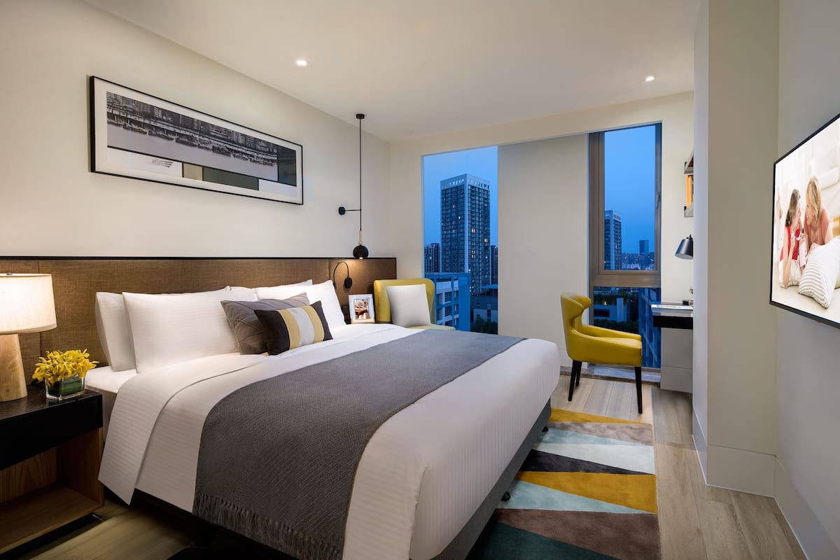 Quarto de hotel moderno com uma cama grande, uma escrivaninha, uma cadeira e vista para a cidade.