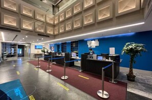 Um lobby de hotel moderno com uma parede azul de destaque, balcão de recepção e uma grande planta em vaso.