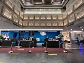 Uma área de recepção de hotel com design moderno, destacando uma parede azul, o balcão de recepção e um painel informativo digital.