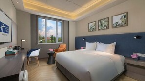 Quarto de hotel com uma cama grande, uma mesa com cadeira, uma mesinha com um vaso e uma janela com vista para o céu e prédios.