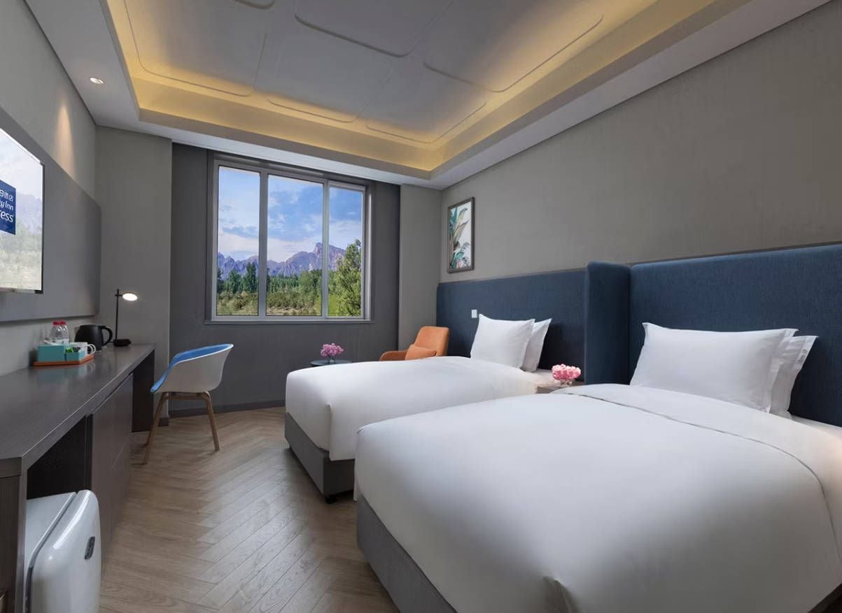 Quarto de hotel com duas camas, uma escrivaninha, uma cadeira e janela com vista para as montanhas.