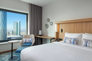 Quarto de hotel com uma cama grande, uma escrivaninha, uma cadeira e vista para a paisagem urbana.